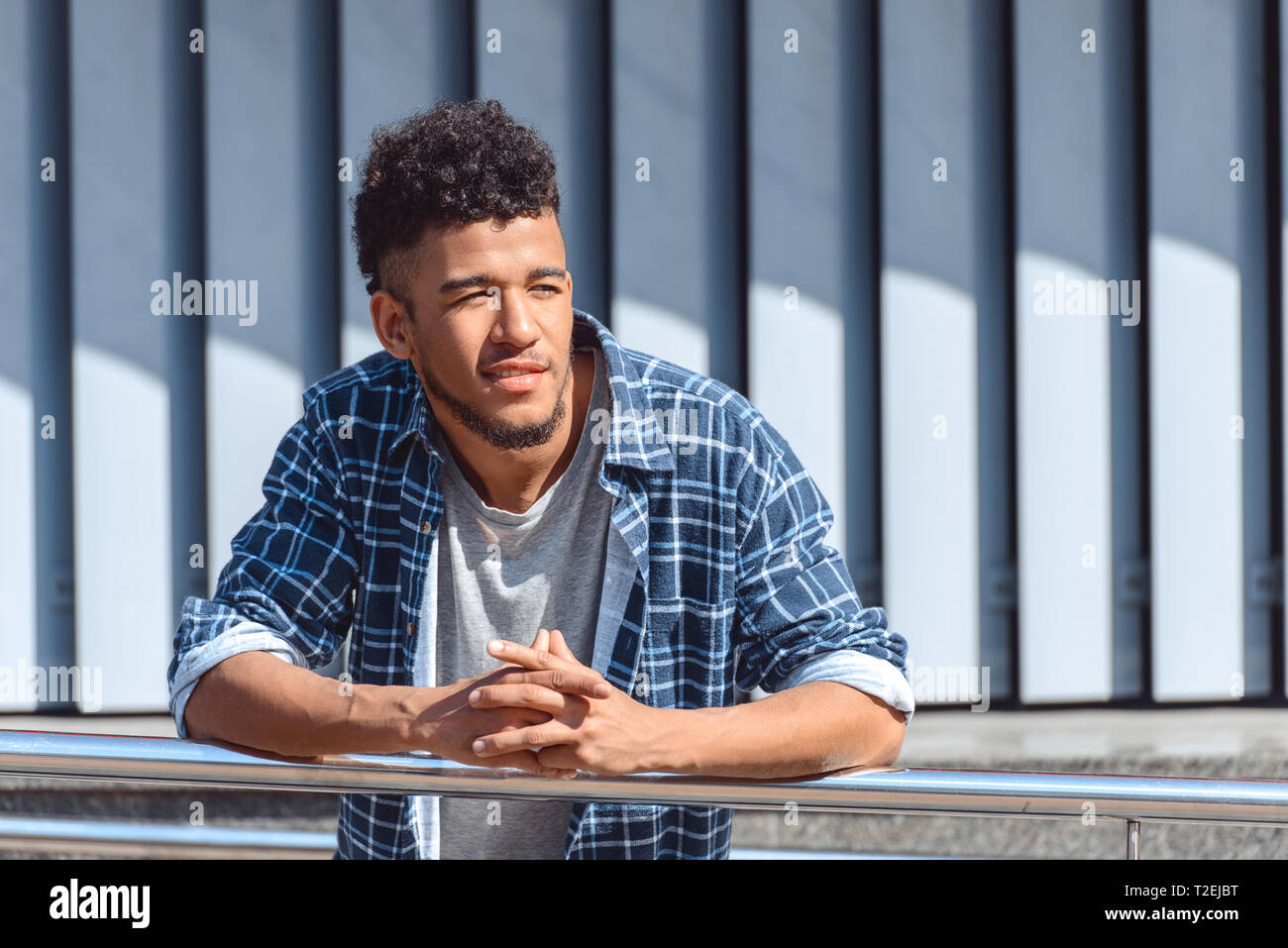 Young mulatto man standing on city street leaning on fence looking ...