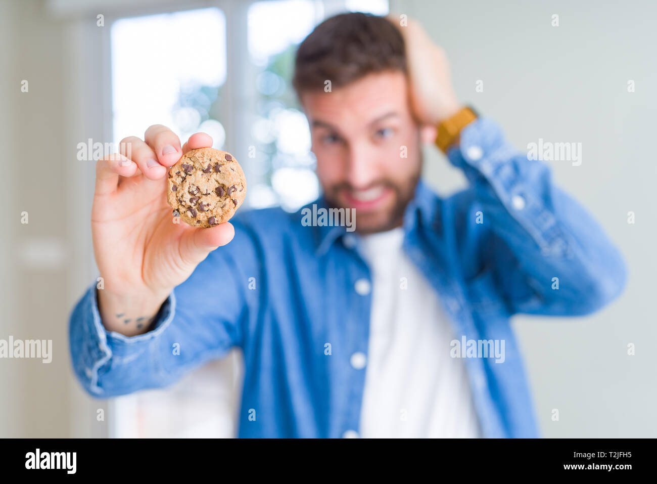 Handsome man eating chocolate chips cookies stressed with hand on head ...