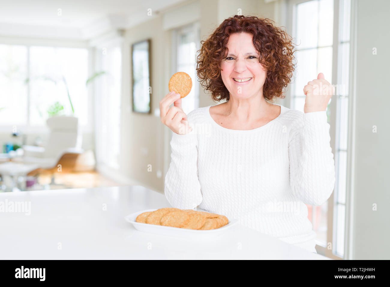 Senior woman eating healthy whole grain biscuit at home screaming proud ...