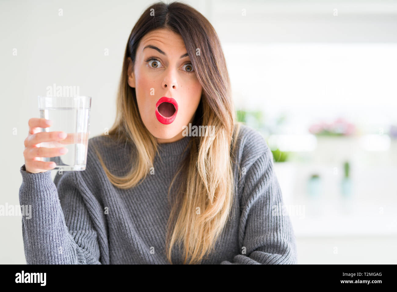 Young beautiful woman drinking glass of water at home scared in shock ...