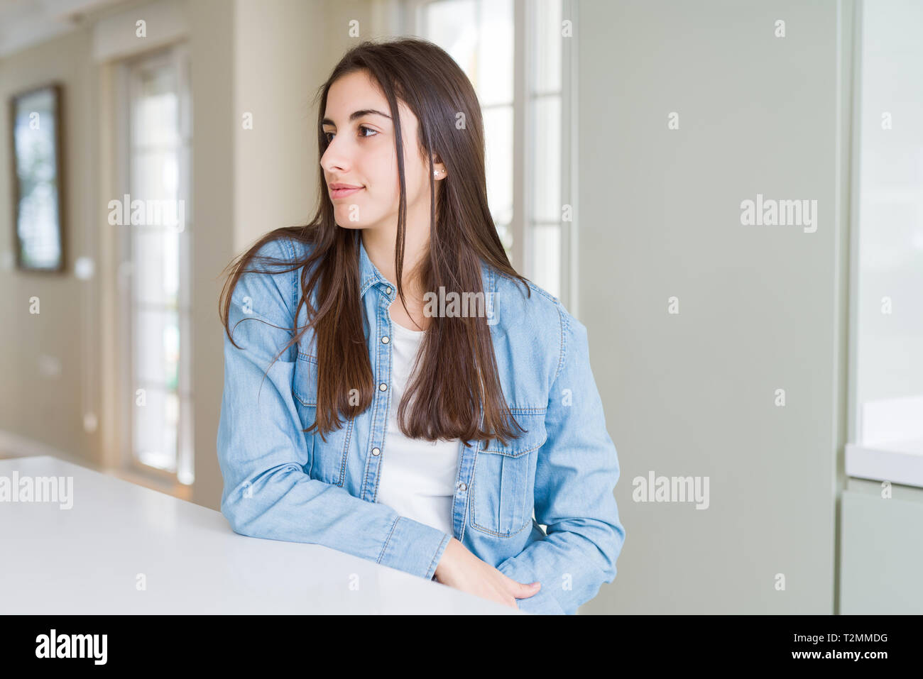 Beautiful young woman sitting on white table at home looking to side ...