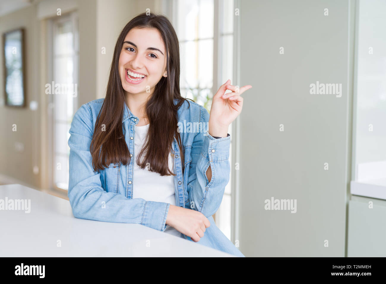 Beautiful young woman sitting on white table at home with a big smile ...