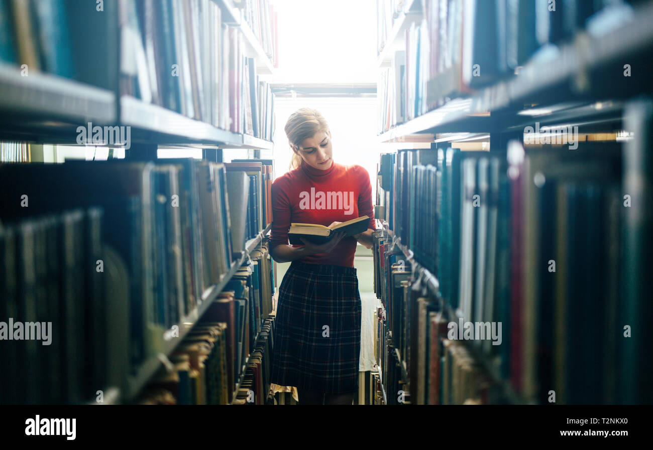 Young attractive librarian reading a book between library bookshelves ...