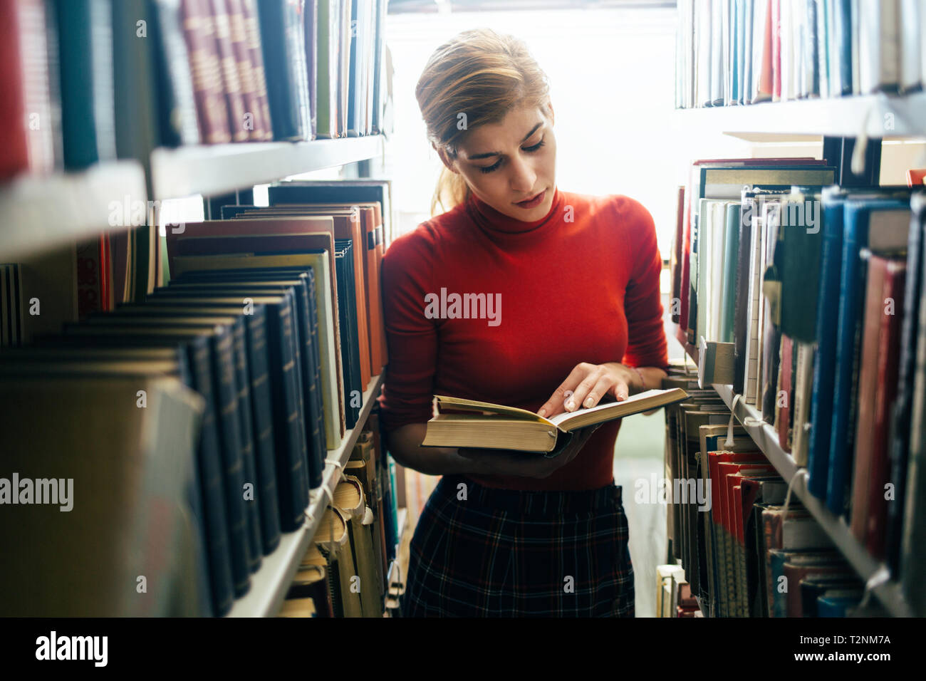 Young attractive librarian reading a book between library bookshelves ...