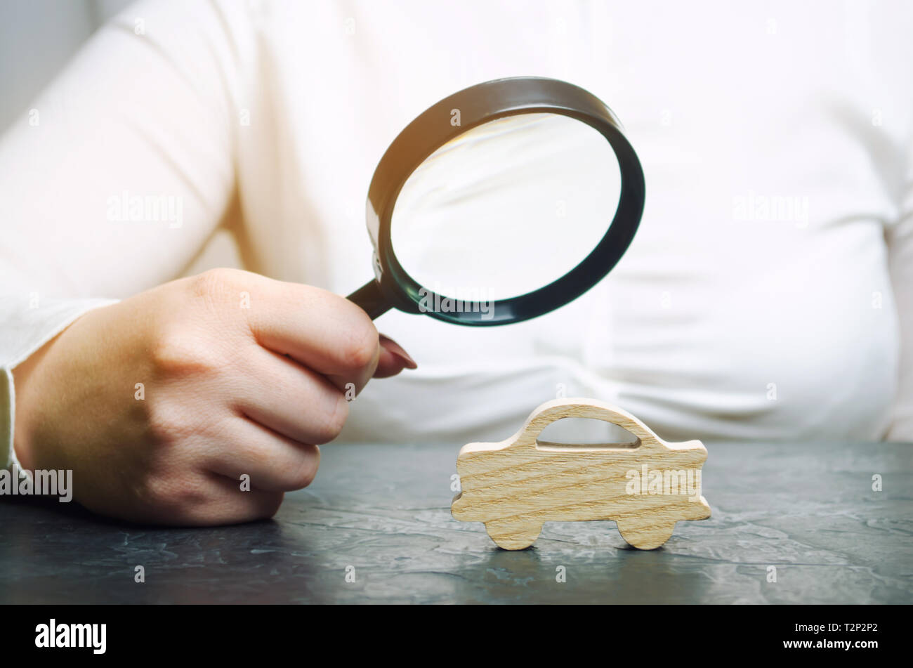 A woman holds a magnifying glass over a miniature wooden car. The ...