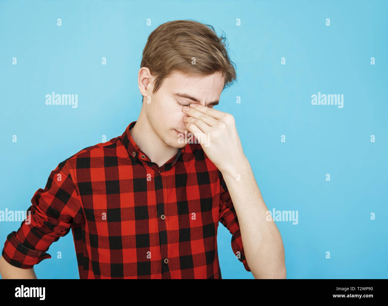 young tired male teenager in red shirt on blue background rubs his eyes ...