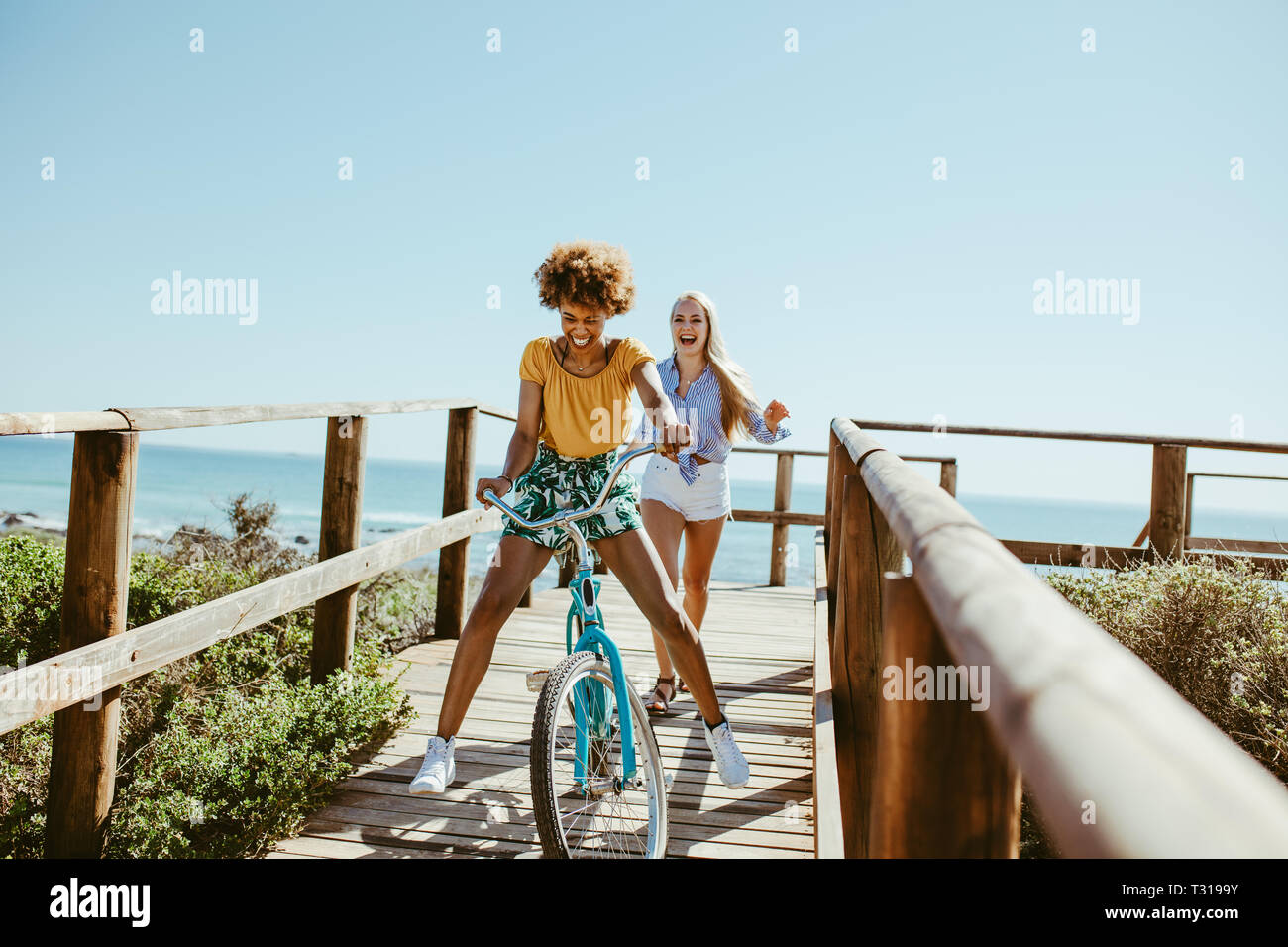 Cheerful woman riding a bicycle on boardwalk with friend running at the ...