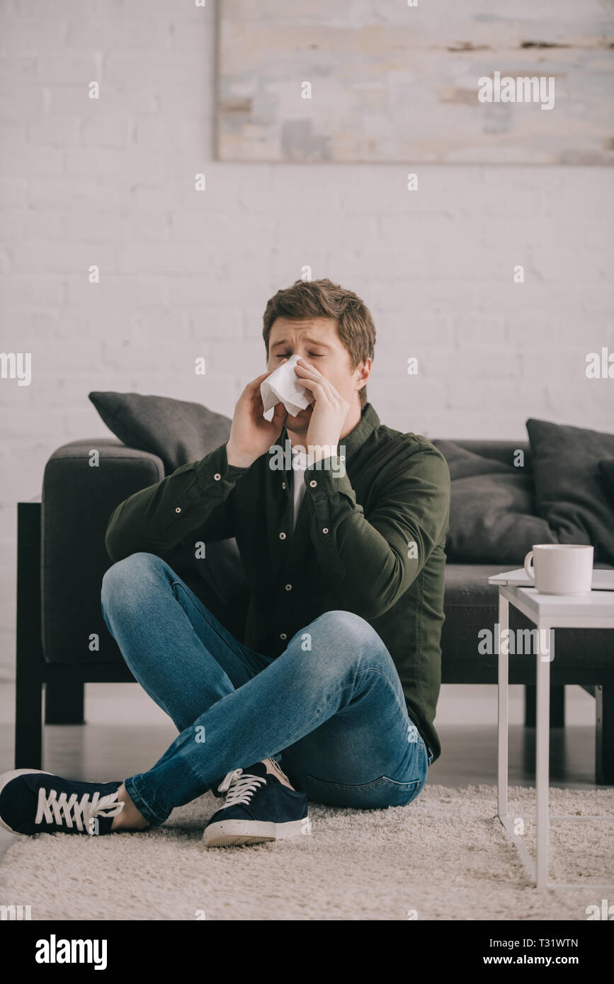 man sitting on carpet with crossed legs and sneezing in tissue near ...