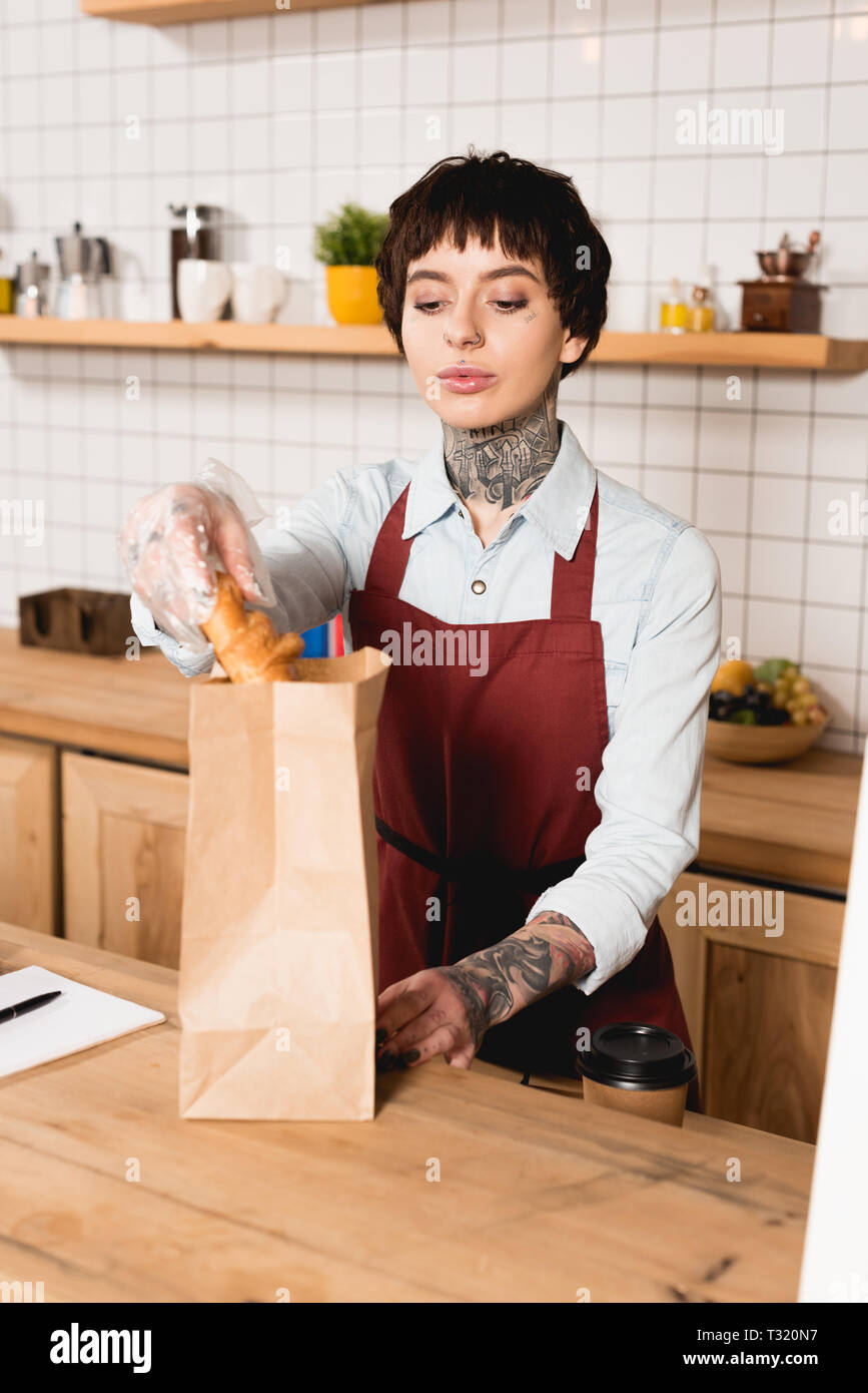 pretty barista in apron putting croissant into paper bag while standing ...