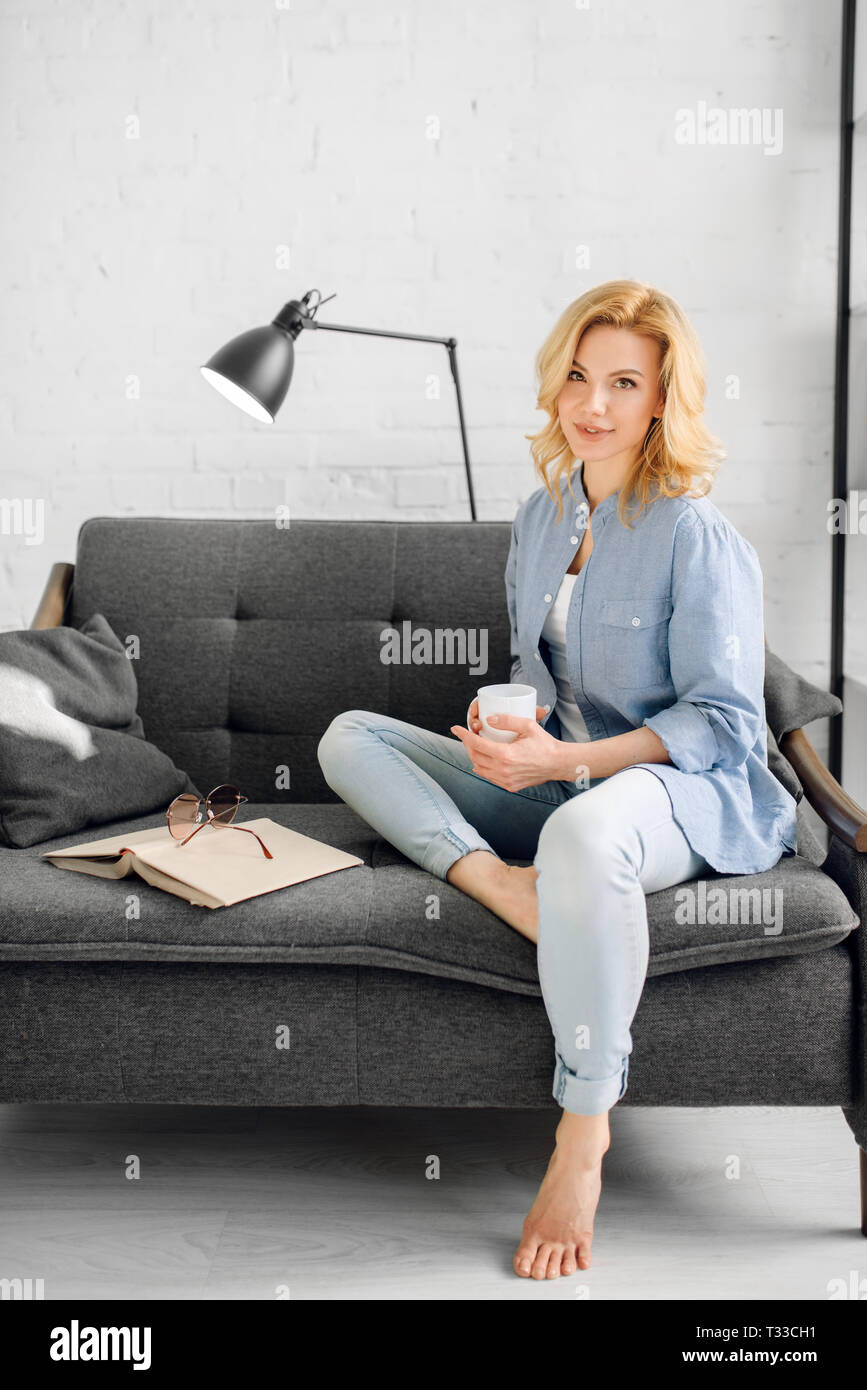 Young lady with book and cup of coffee poses on cozy black couch ...