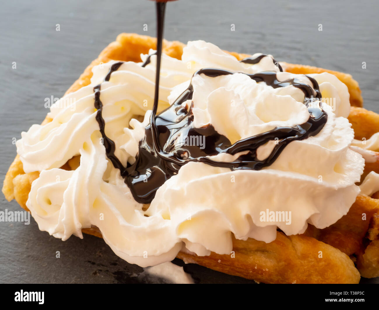 Chocolate syrup falling on a Belgian waffle with cream on a slate plate ...