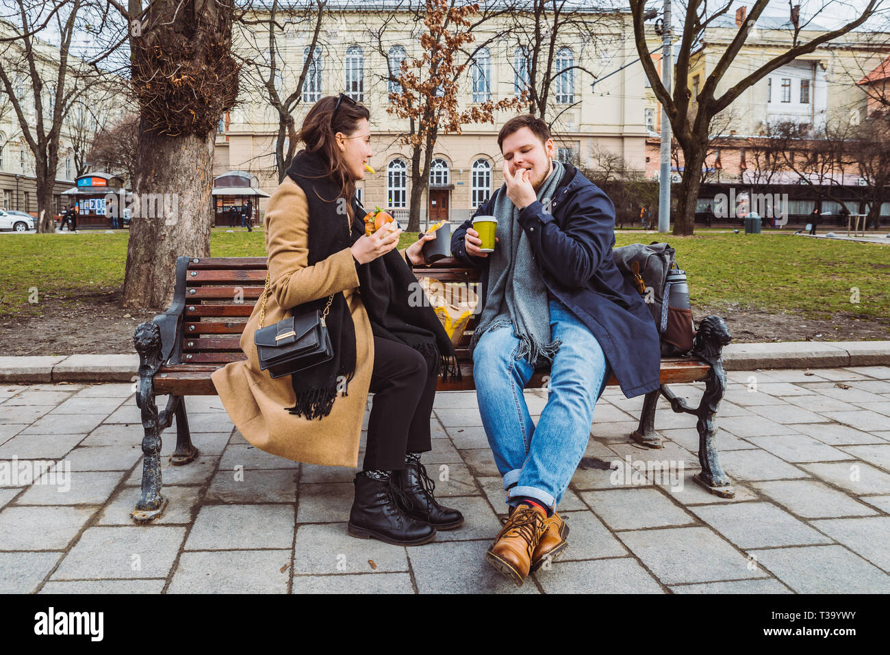 young adult couple eating fast food on bench of city park. lifestyle ...