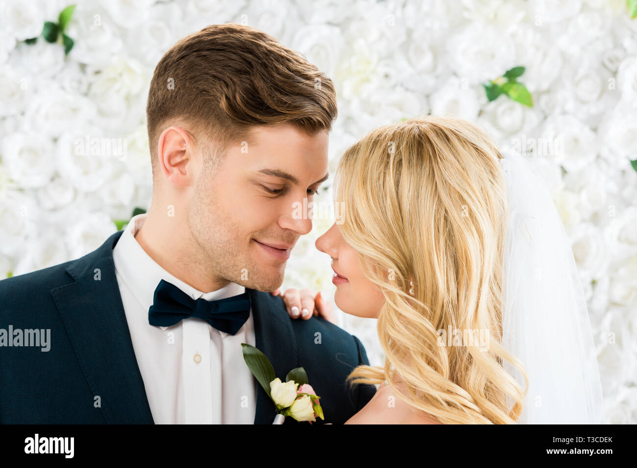 happy young groom and bride standing face to face on white floral ...