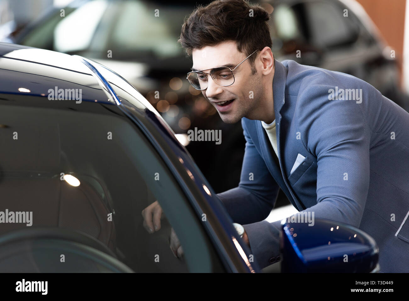 happy customer in glasses standing near automobile in car showroom ac ...