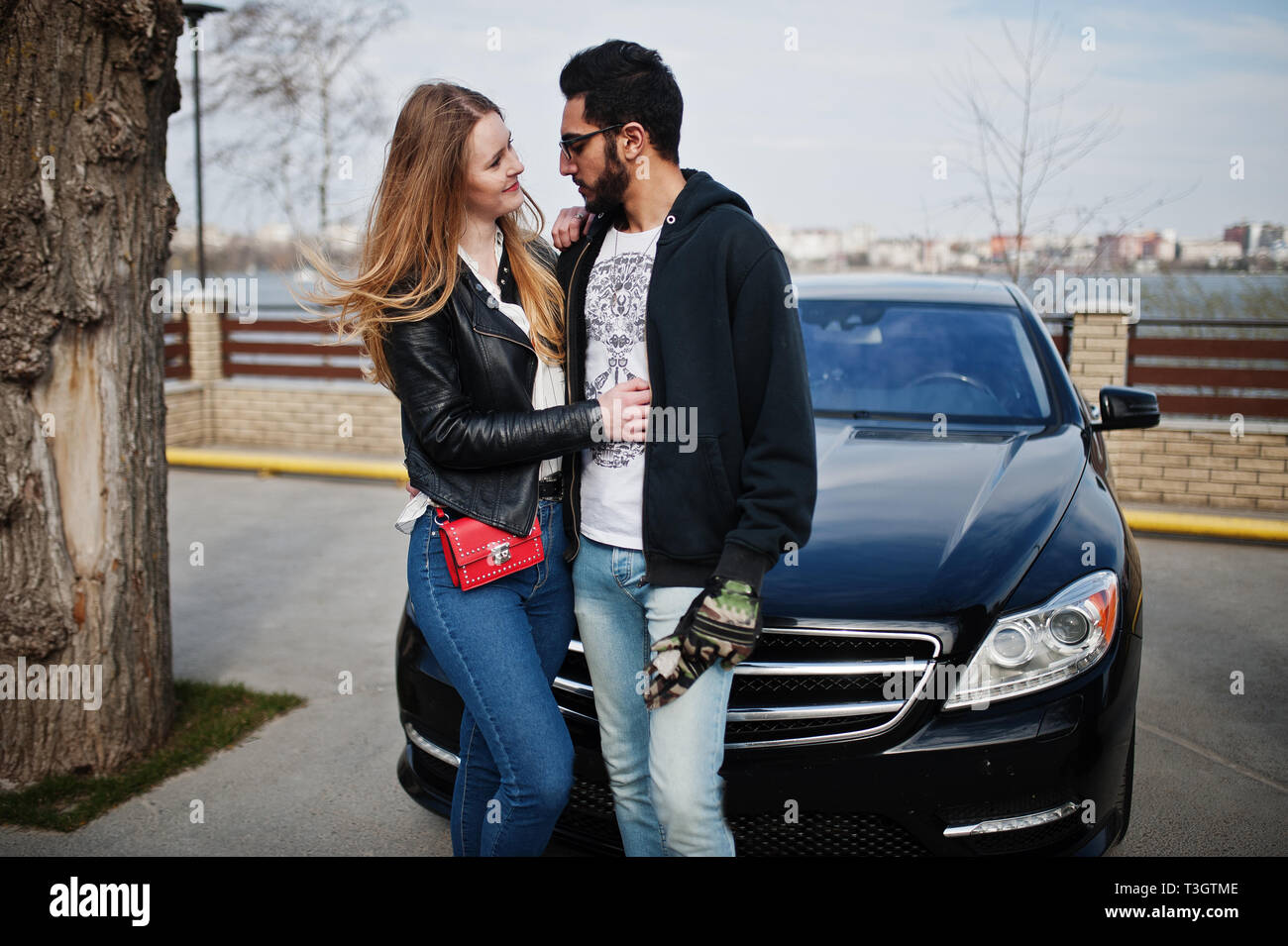 Cool multiracial couple hugs together against black elegant sport car ...