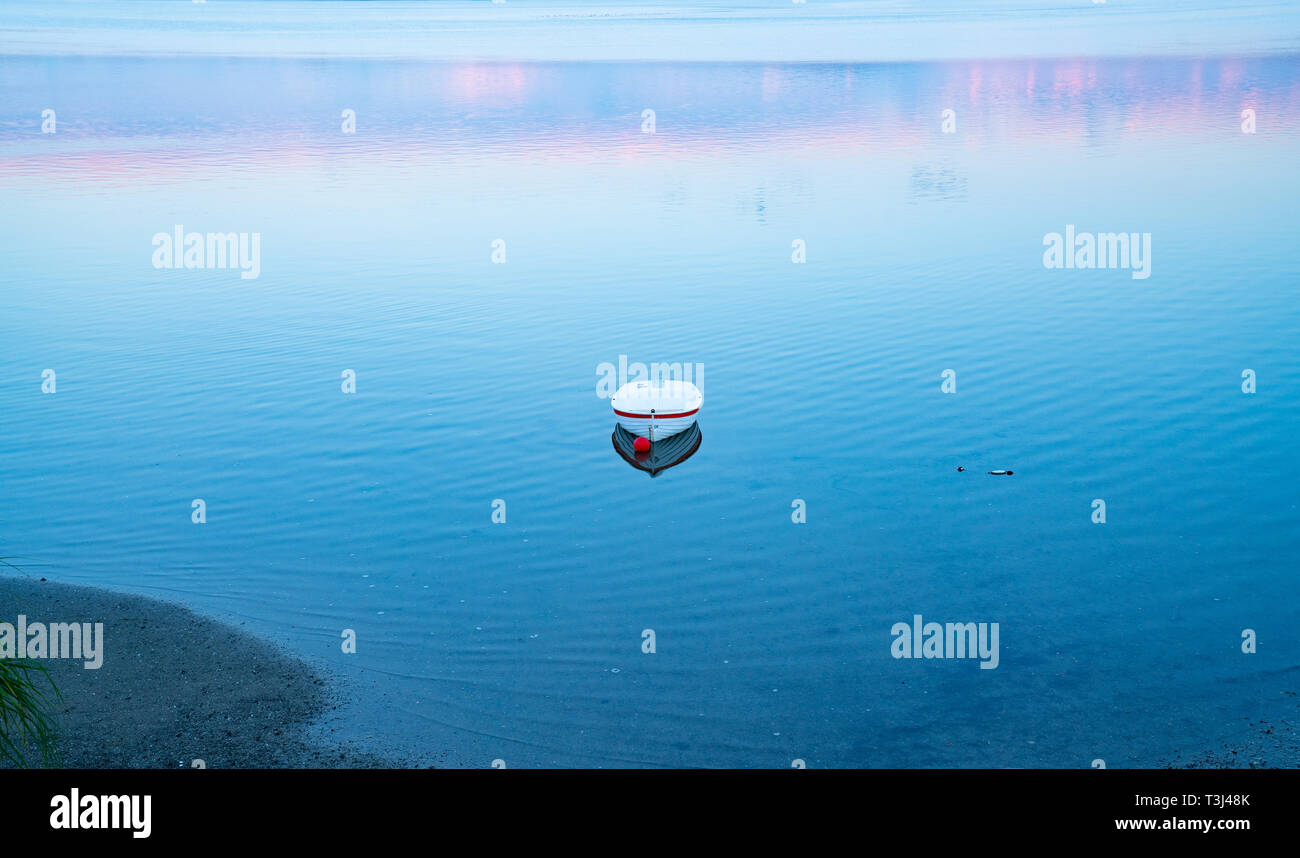 White dinghy afloat on calm blue water with reflection of sunset colors ...