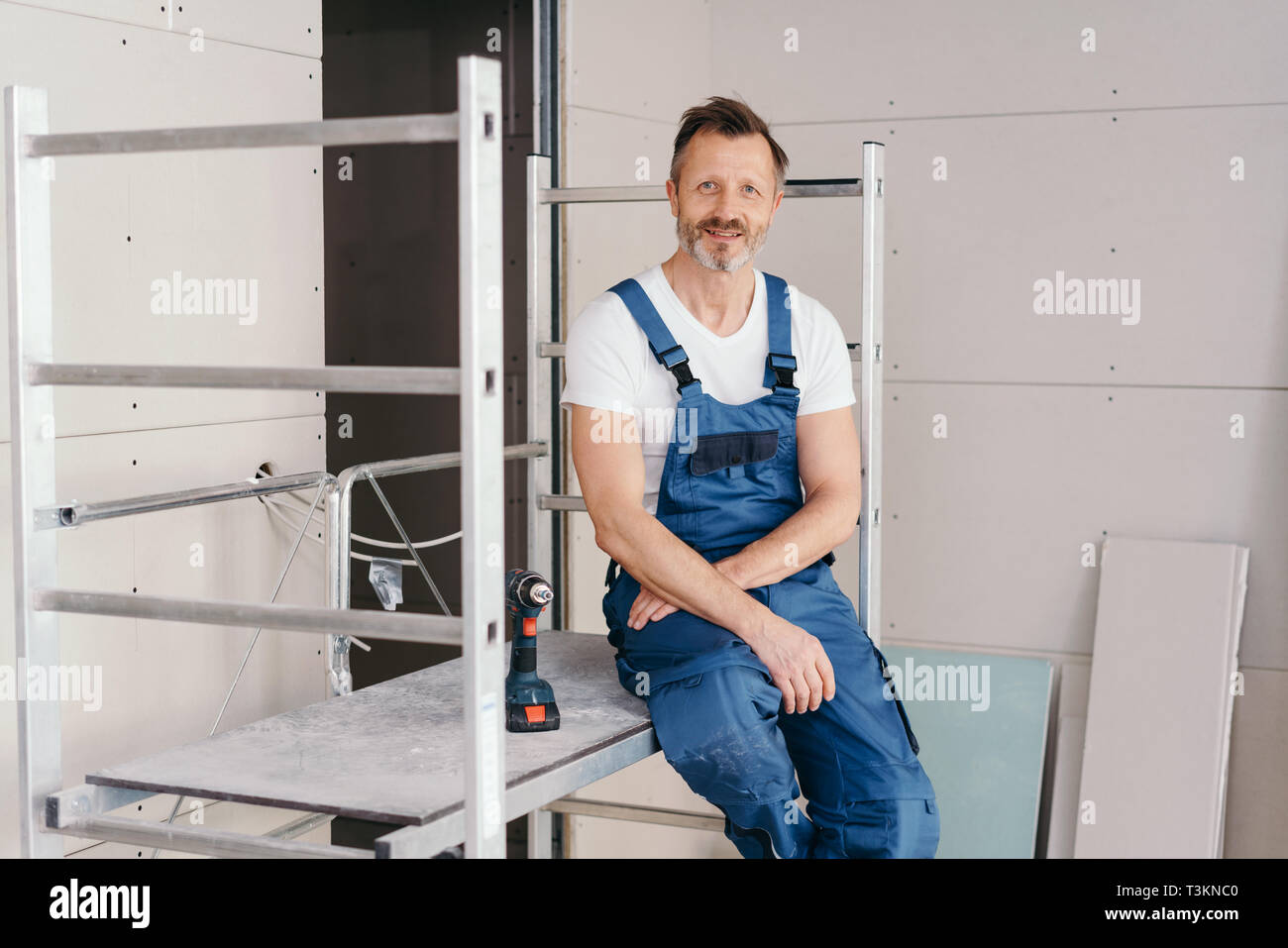 Builder in denim overalls relaxing on low scaffolding indoors in a new ...