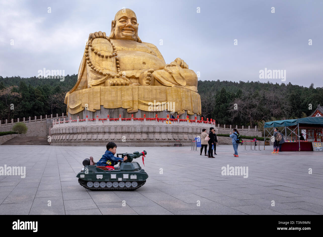 Child driving a miniature tank in front of big golden statue of Buddha ...