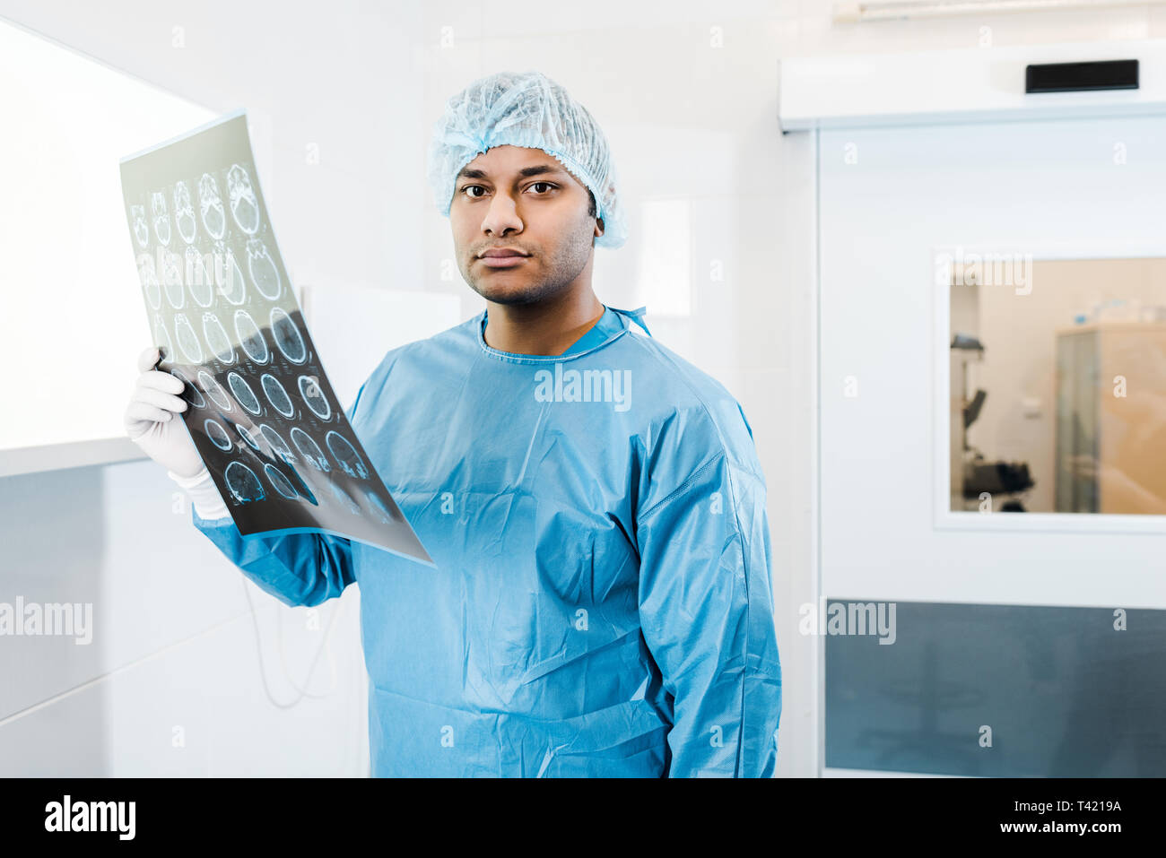 handsome doctor in medical cap and uniform holding x-ray and looking at ...