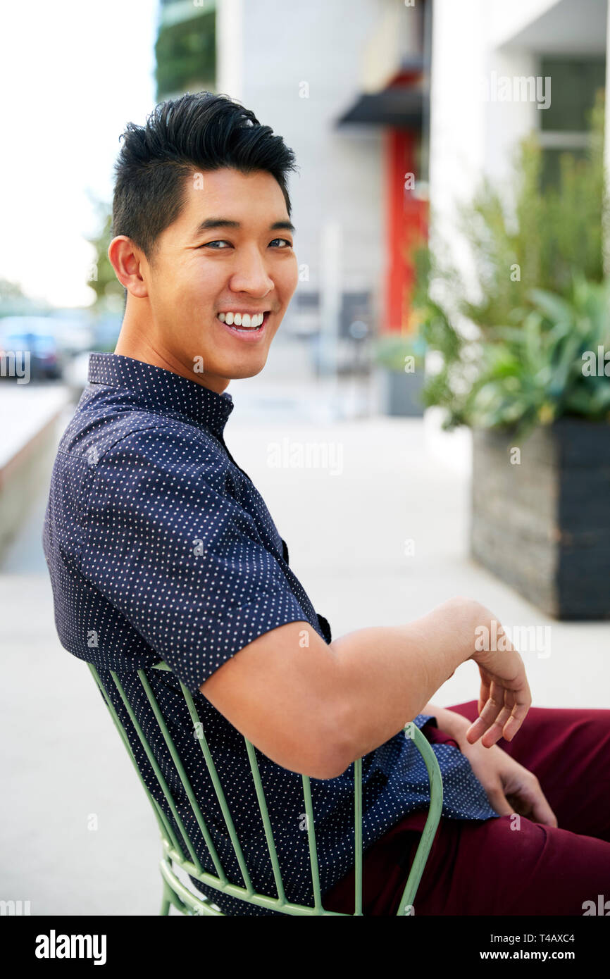 Millennial Asian man sitting on a chair in a city street, turning and ...