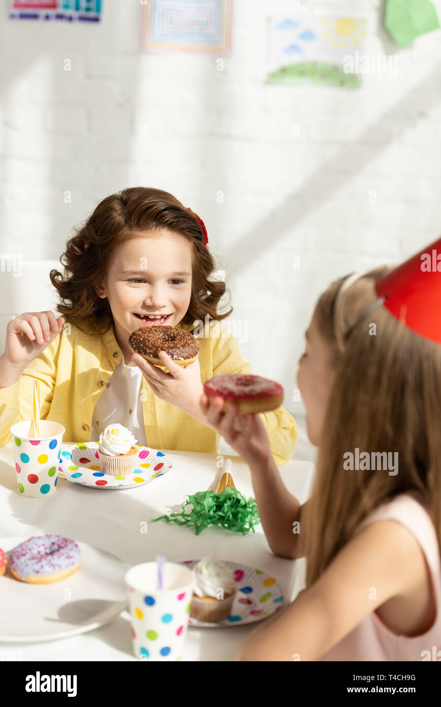 adorable kids sitting at party table and eating donuts during birthday ...
