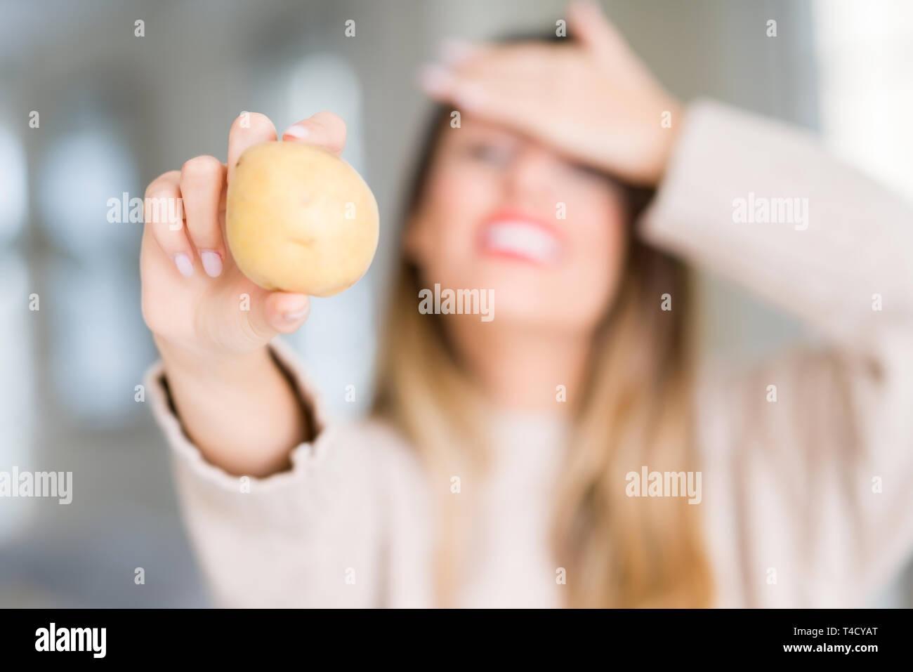 Young beautiful woman holding fresh potato at home stressed with hand ...