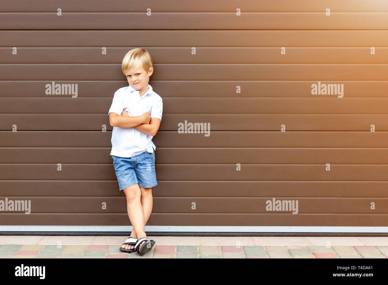 Cute grumpy blond child in casual clothing standing against brown ...