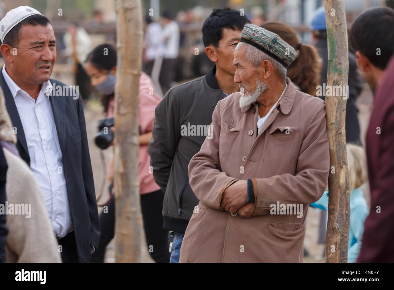 Bearded Uyghur man with a doppa (traditional hat) leaning at a pole at ...