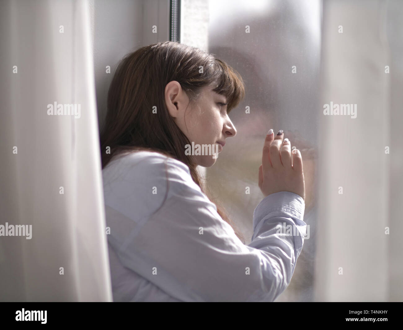 Sad brunette woman looks out the window with her hand on the glass ...