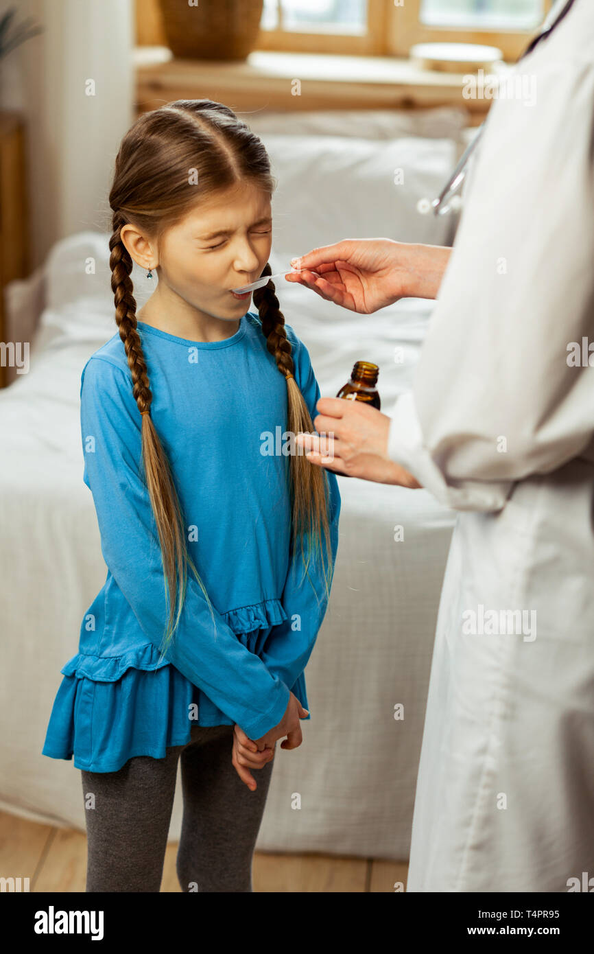 A girl taking syrup. Small pretty sick brown-haired girl with braids ...
