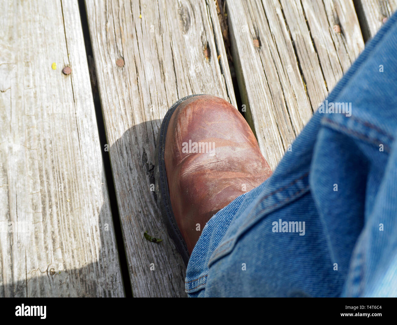 A scuffed brown leather boot with blue jeans on weathered wooden deck ...