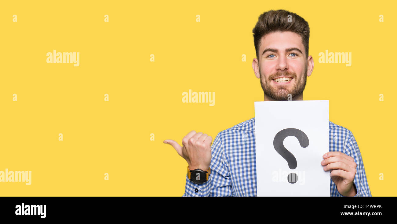 Young handsome man holding paper with question mark pointing and ...