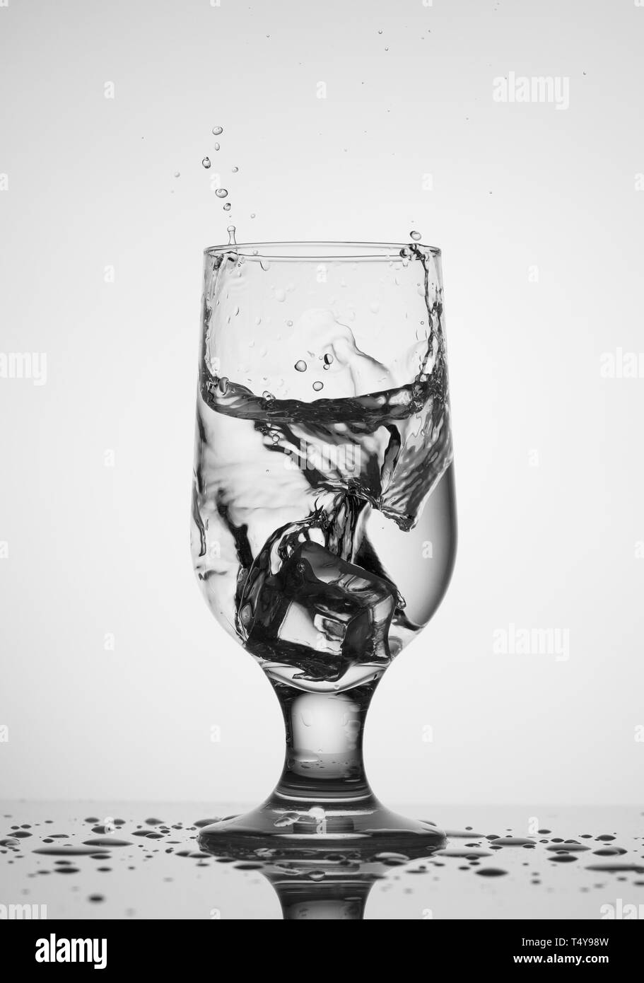 The cube of ice falls into a clear glass glass with water. Splash of small drops of water on the ...
