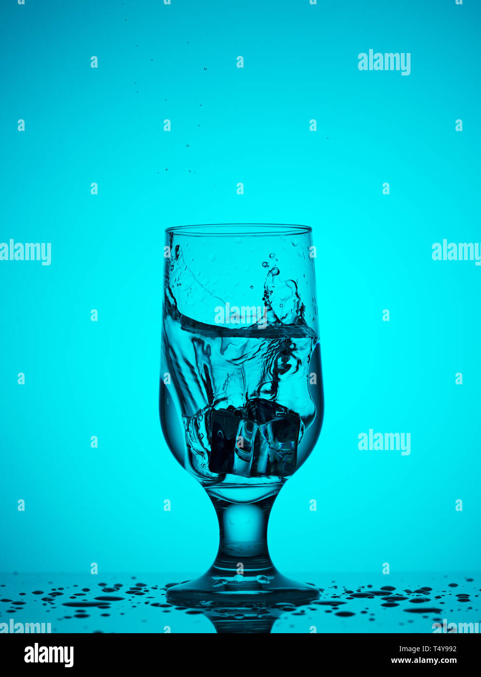 Splash of water from a falling ice cube into a transparent glass glass on a turquoise background ...