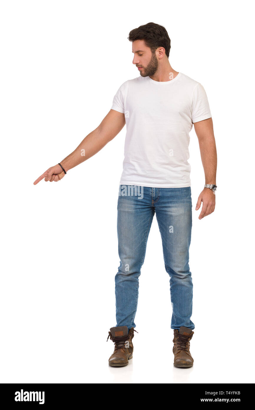 Serious young man in jeans and white t-shirt is standing, pointing down ...