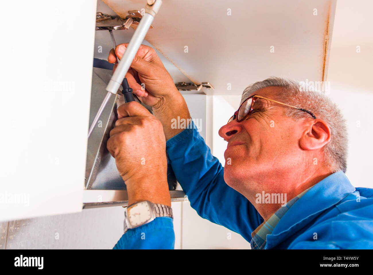 Senior workman is fixing the cupboard in the kitchen with screwdriver ...