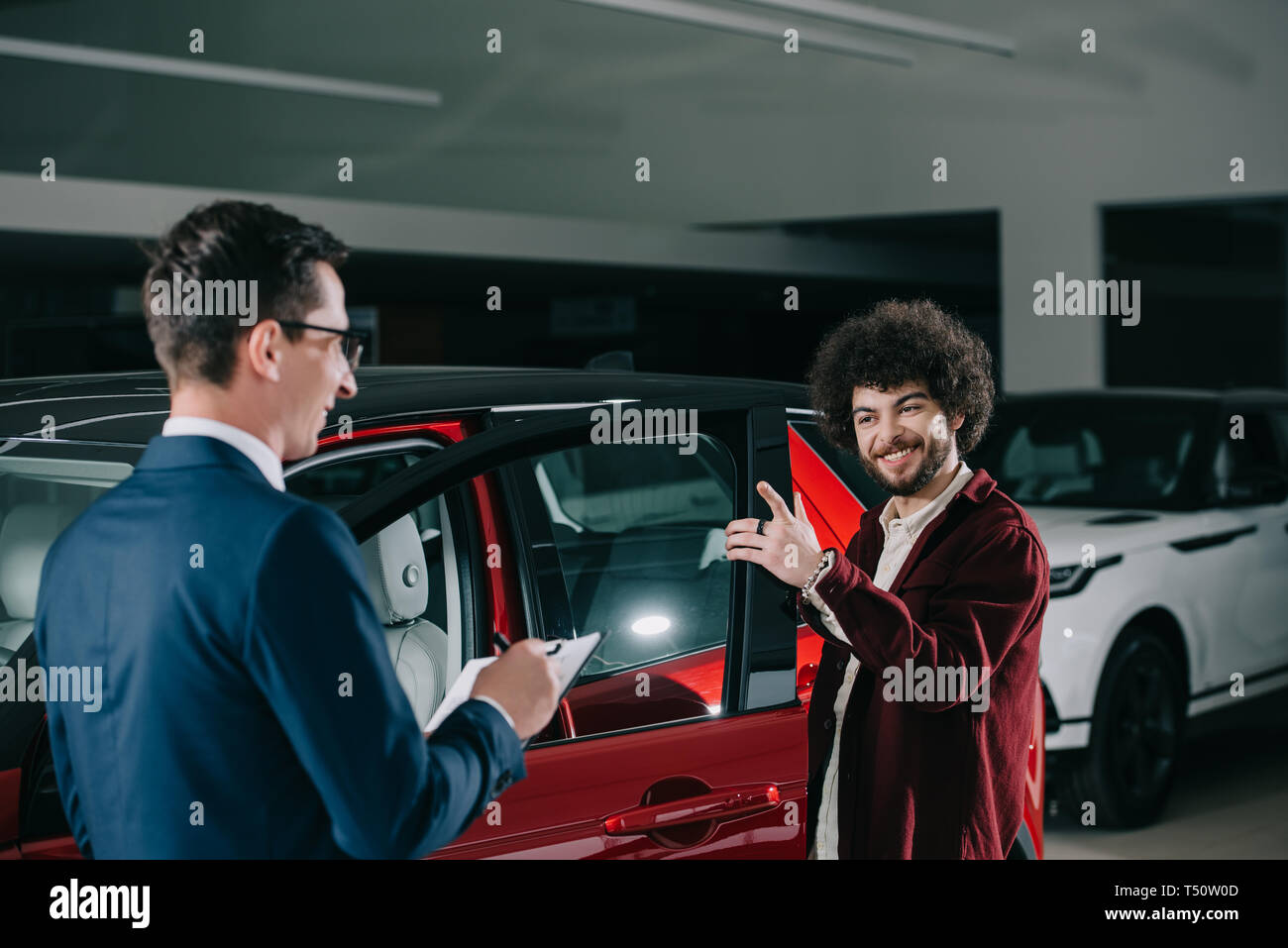 cheerful curly man looking at car dealer in glasses while standing near ...