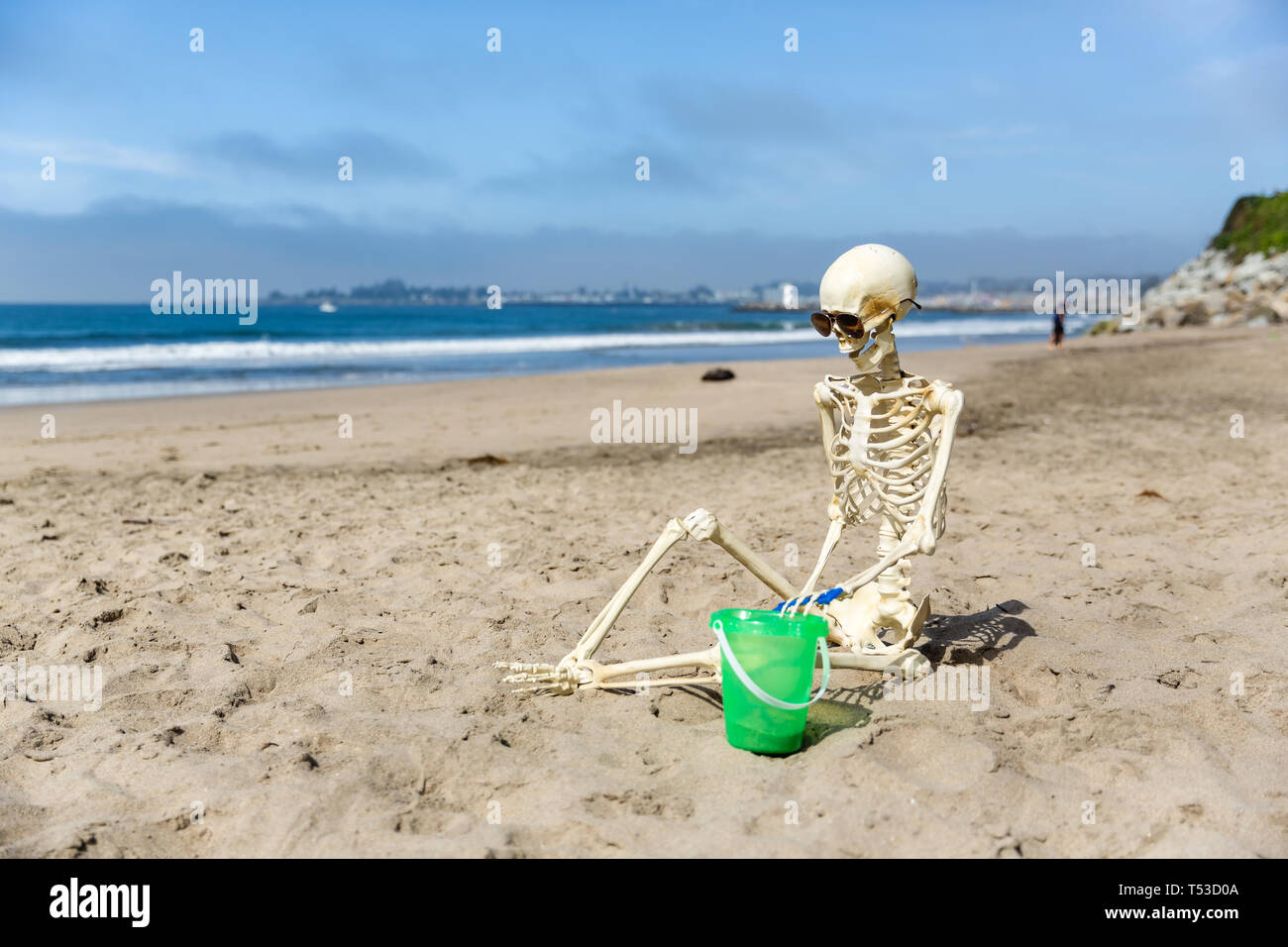 Skeleton sits on the beach playing in the sand with a bucket and shovel ...