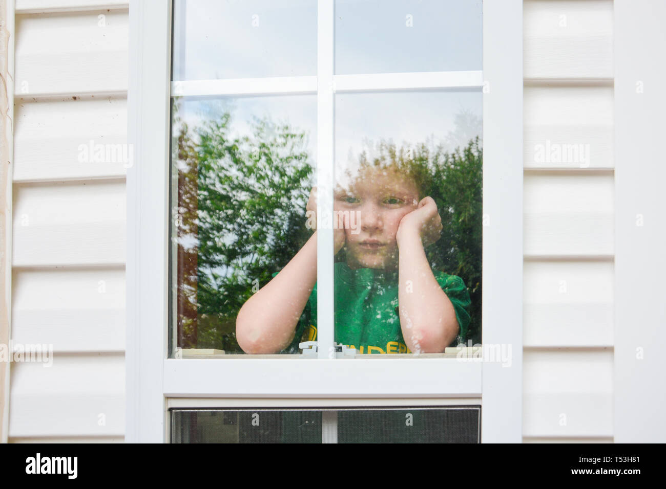 A young boy is stuck alone indoors and sad gazes out the window at the ...