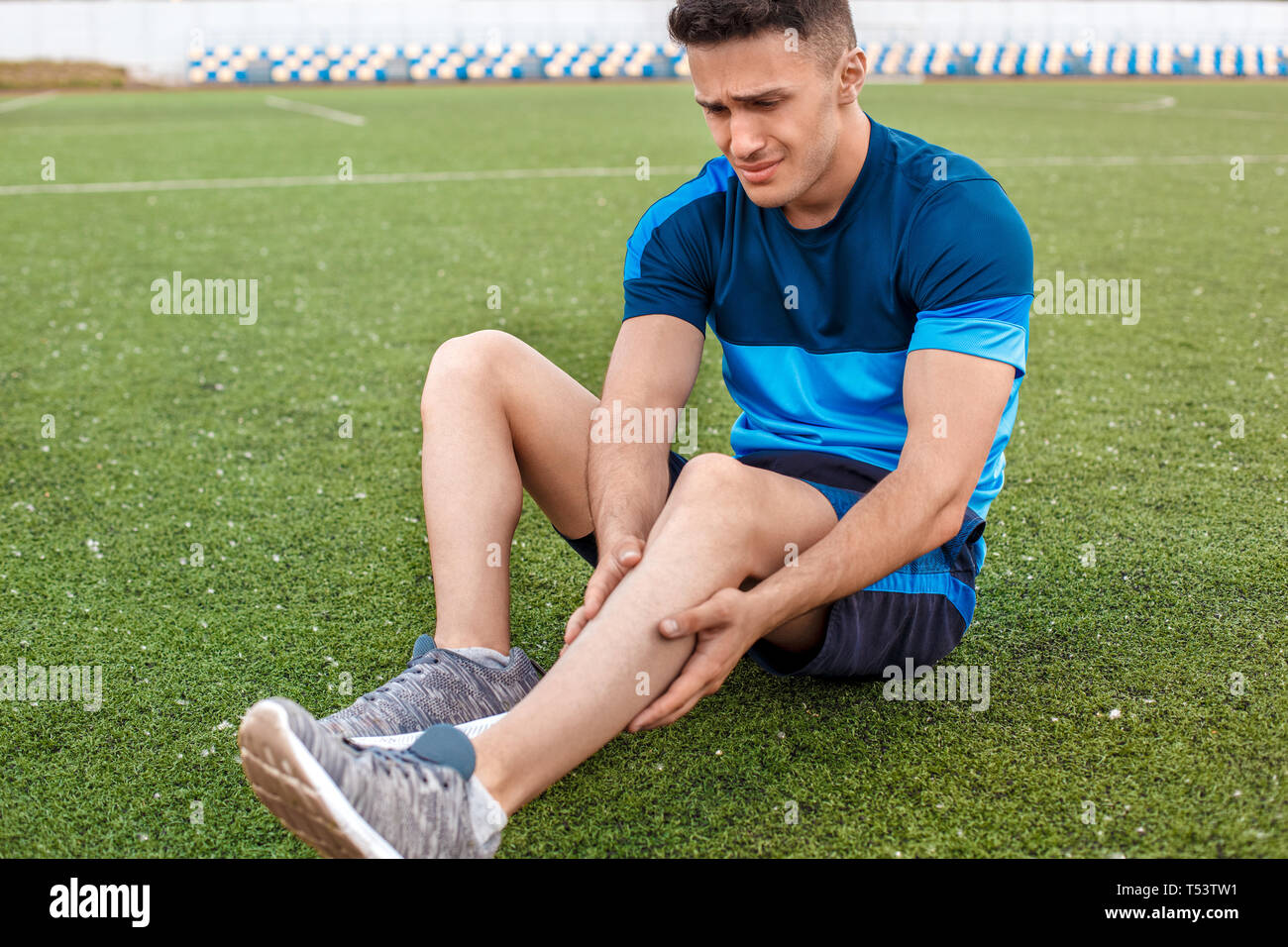 Young sportsman on stadium outside sitting on lawn hurted leg muscle ...