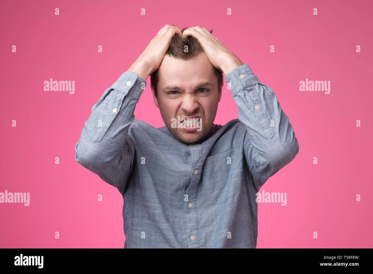Angry screaming young man putting hands on head on pink wall in studio ...