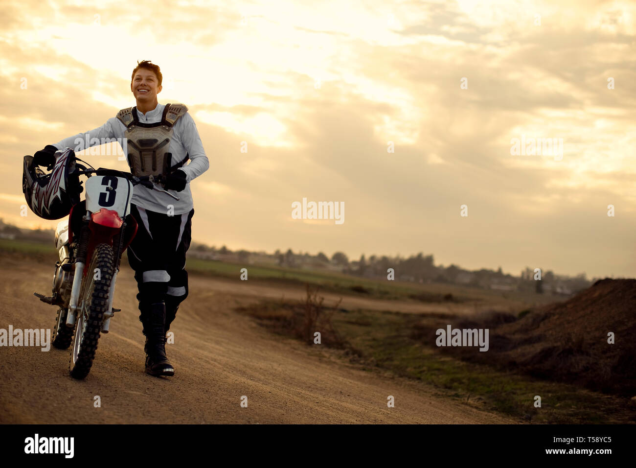 Smiling young female motocross rider pushing her dirt bike at the track ...