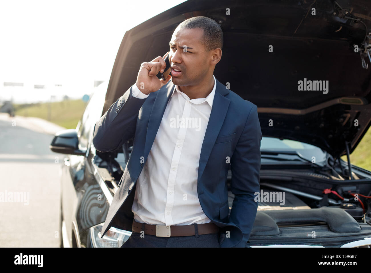Young african american man leaning on car with opened hood talking on ...