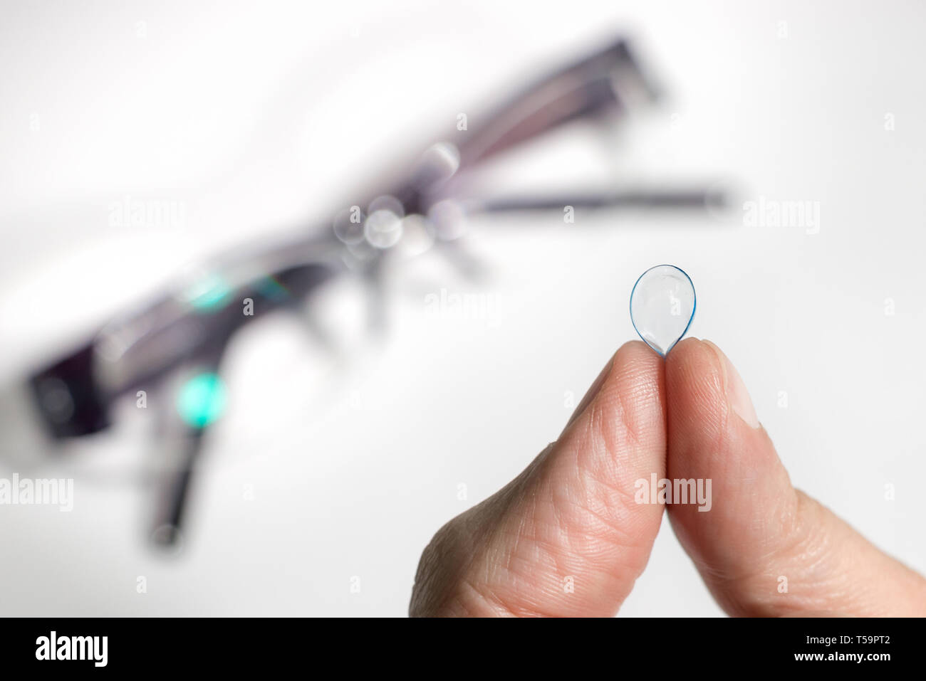 A male finger holding a contact eye lens over blurred eyeglasses as ...