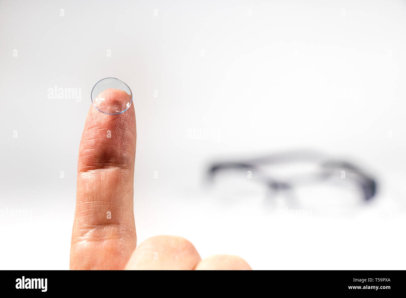 A male finger holding a contact eye lens over blurred eyeglasses as ...