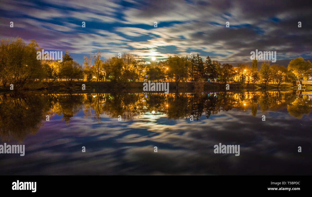 Night landscape above river with trees lit by city lights and clouds in ...
