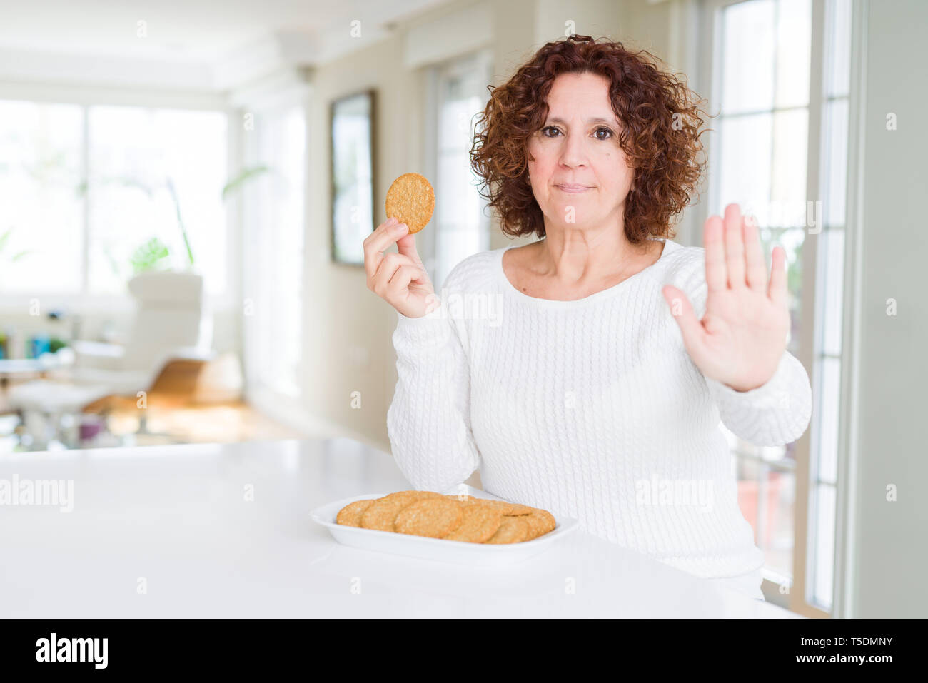 Senior woman eating healthy whole grain biscuit at home with open hand ...