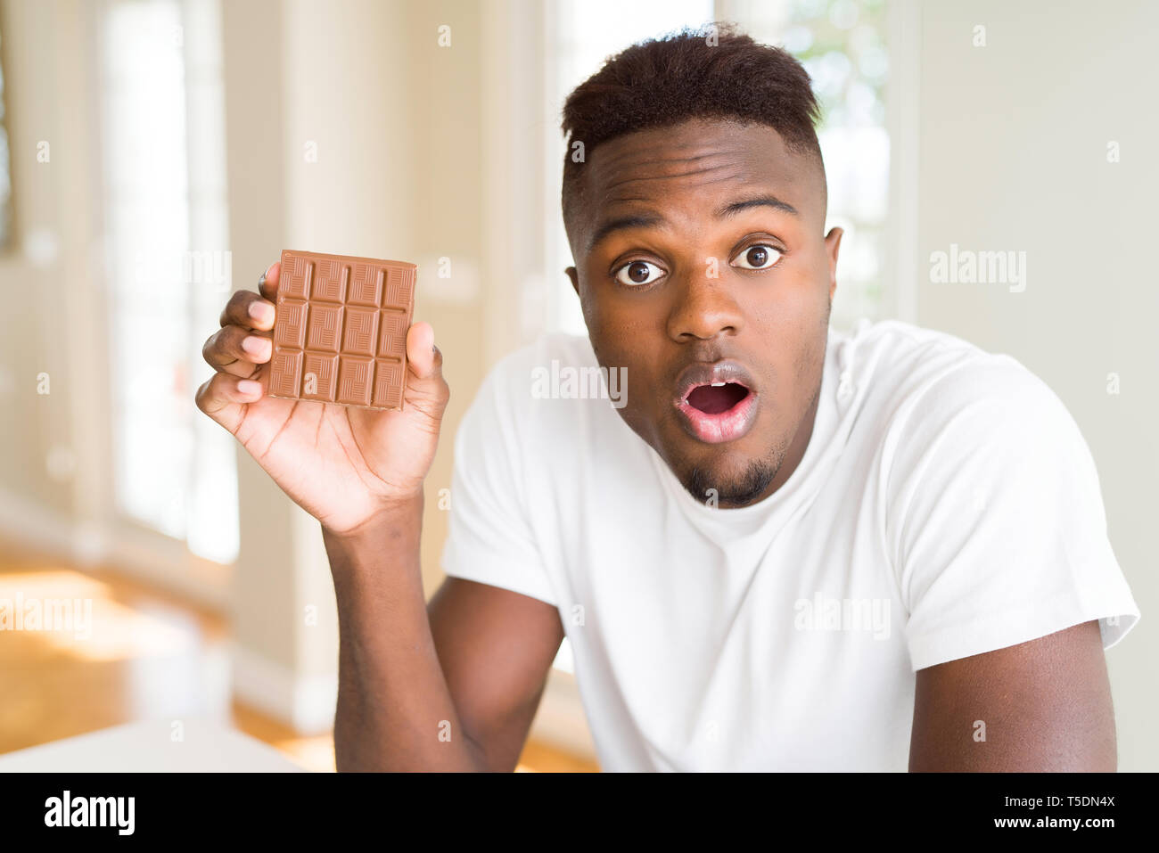 Young african american man eating chocolate bar scared in shock with a ...