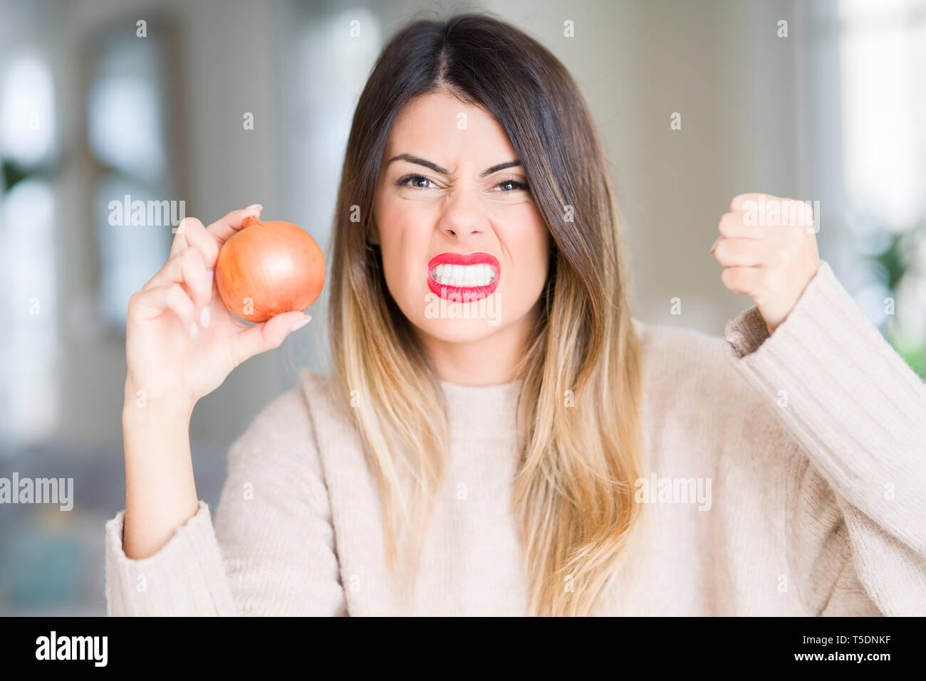 Young beautiful woman holding fresh onion at home annoyed and ...