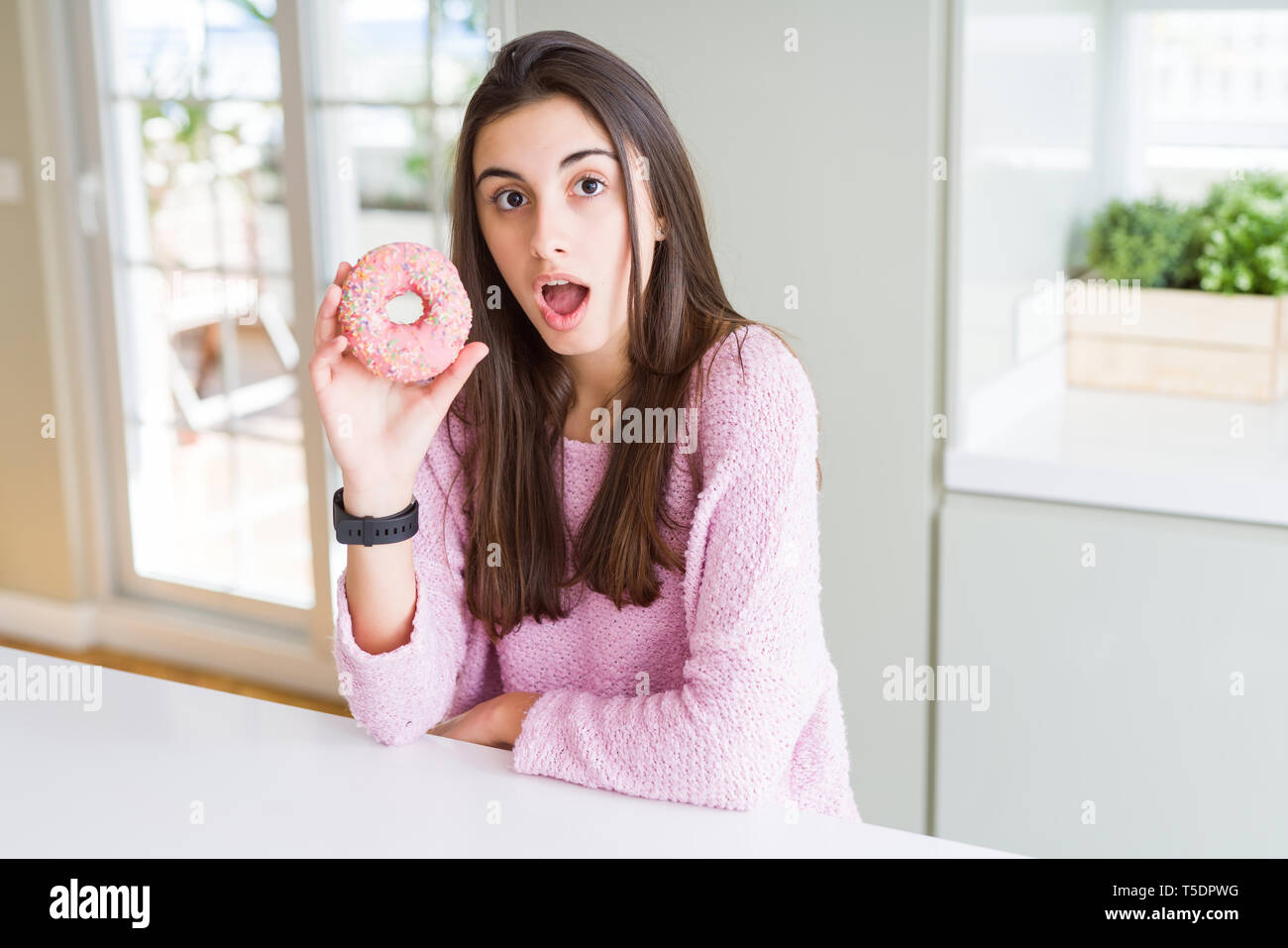 Beautiful young woman eating pink chocolate chips donut scared in shock ...